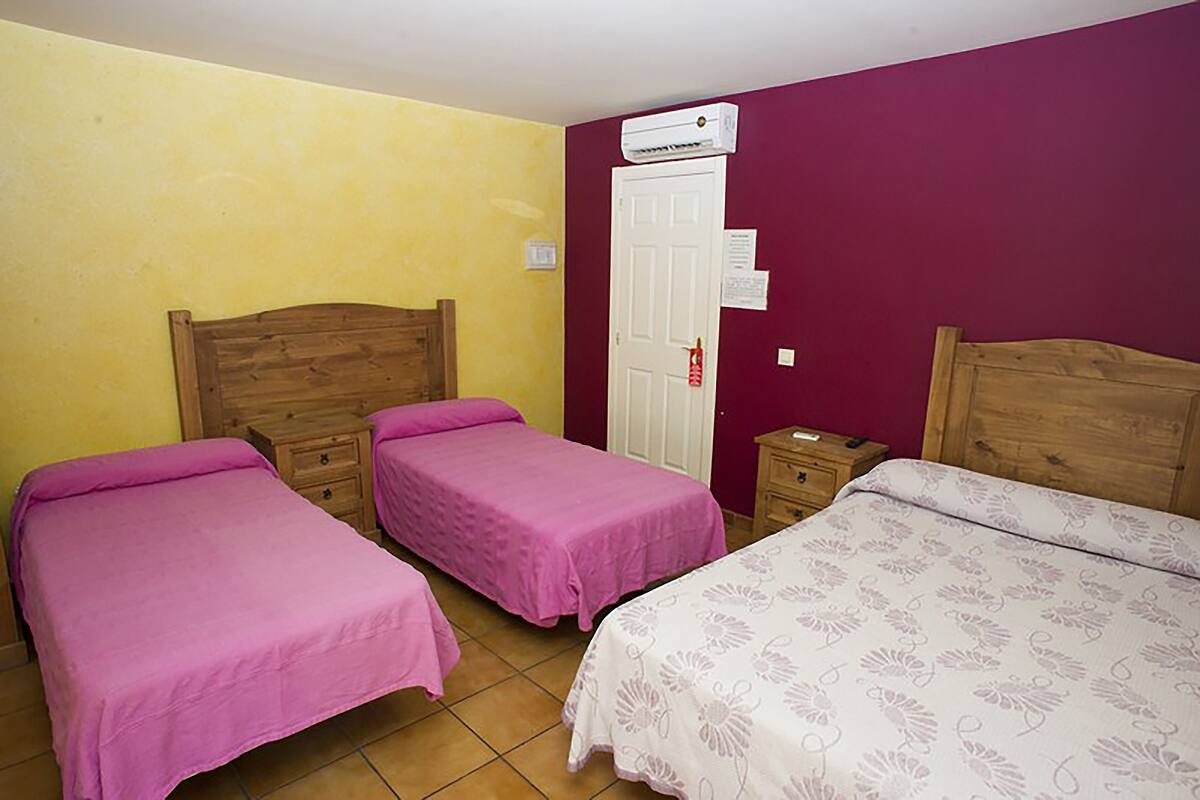 Quarto com três camas, cada uma com roupa de cama diferente. Há uma porta branca com um extintor fixado, e um aparelho de ar condicionado acima da porta.