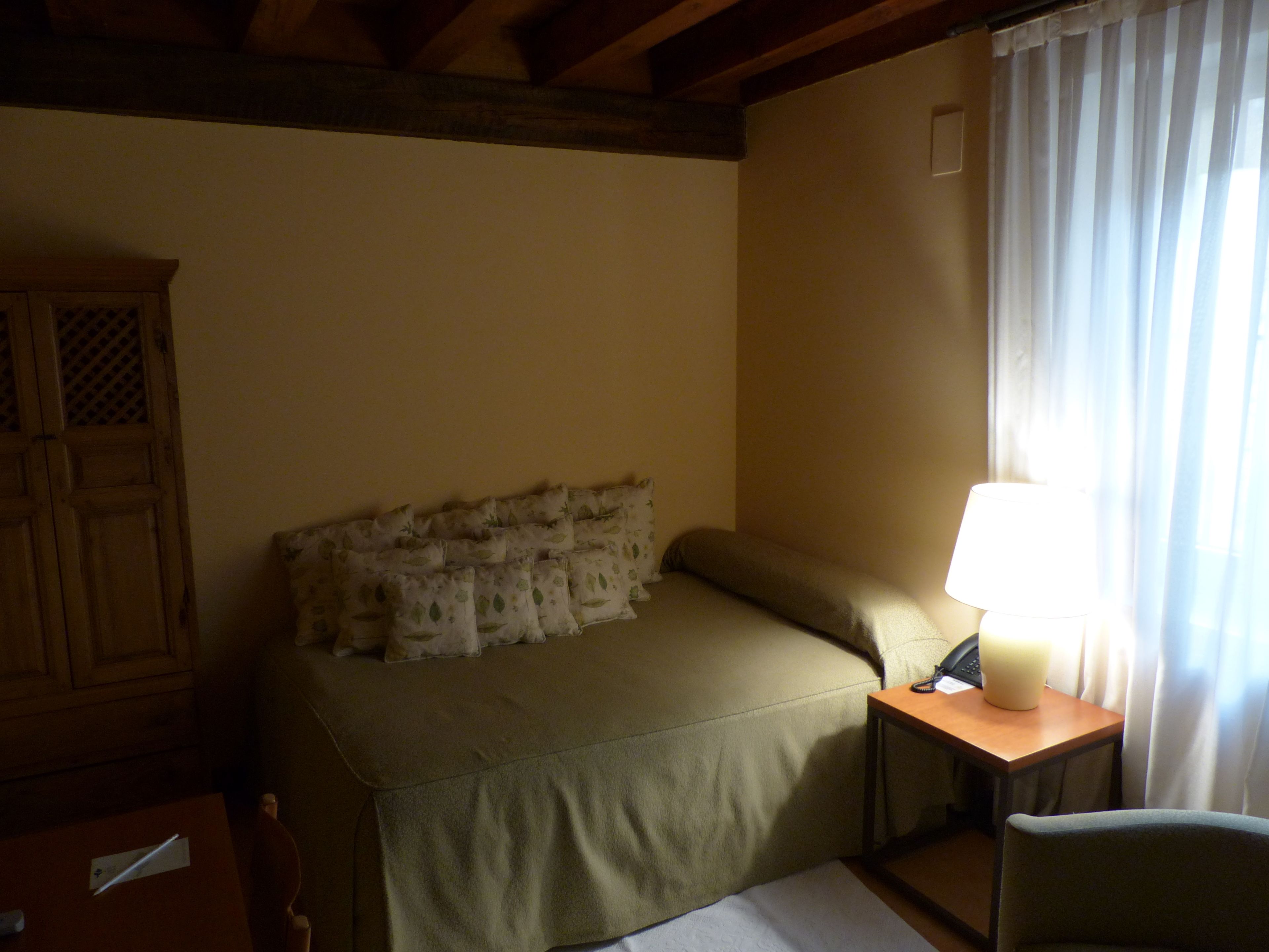 Uma cama de solteiro com travesseiro estampado, mesa de cabeceira com abajur, uma cadeira e um guarda-roupa de madeira.
