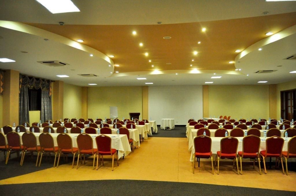 Sala de conferências com mesas e cadeiras dispostas em fileiras, equipada com projetores e garrafas de água.