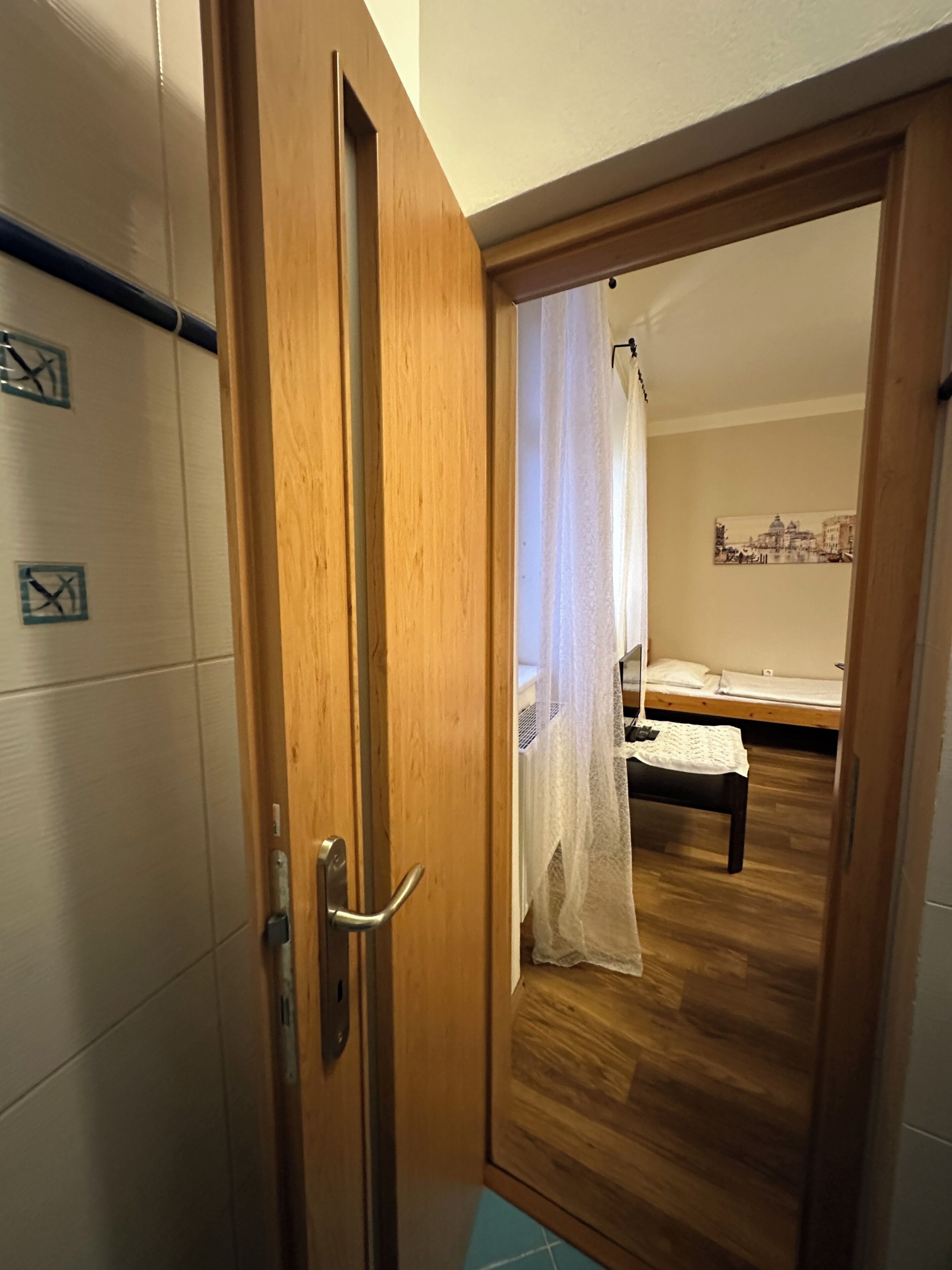 Uma porta de madeira que dá para um quarto com uma cama, uma cortina branca e um quadro na parede.