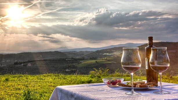 Uma mesa com uma garrafa e copos de vinho, uma taça de morangos e um prato de pão e queijo, com uma paisagem montanhosa ao fundo.