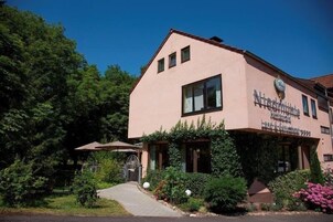 Um edifício de dois andares com uma placa que diz "Niestracht Hotel". Há uma calçada que leva à entrada, e o edifício é cercado por vegetação.