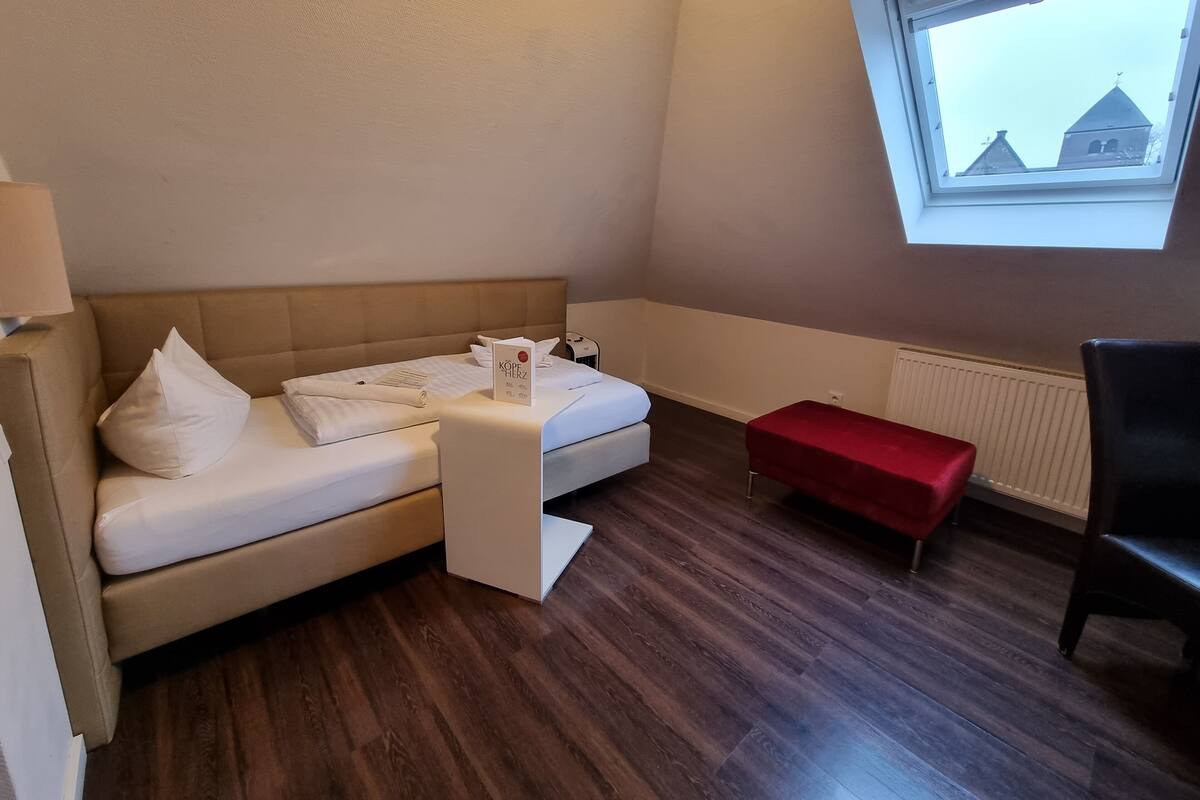 Quarto pequeno e bem iluminado com uma cama, um banquinho vermelho, uma janela com vista para edifícios e um radiador.