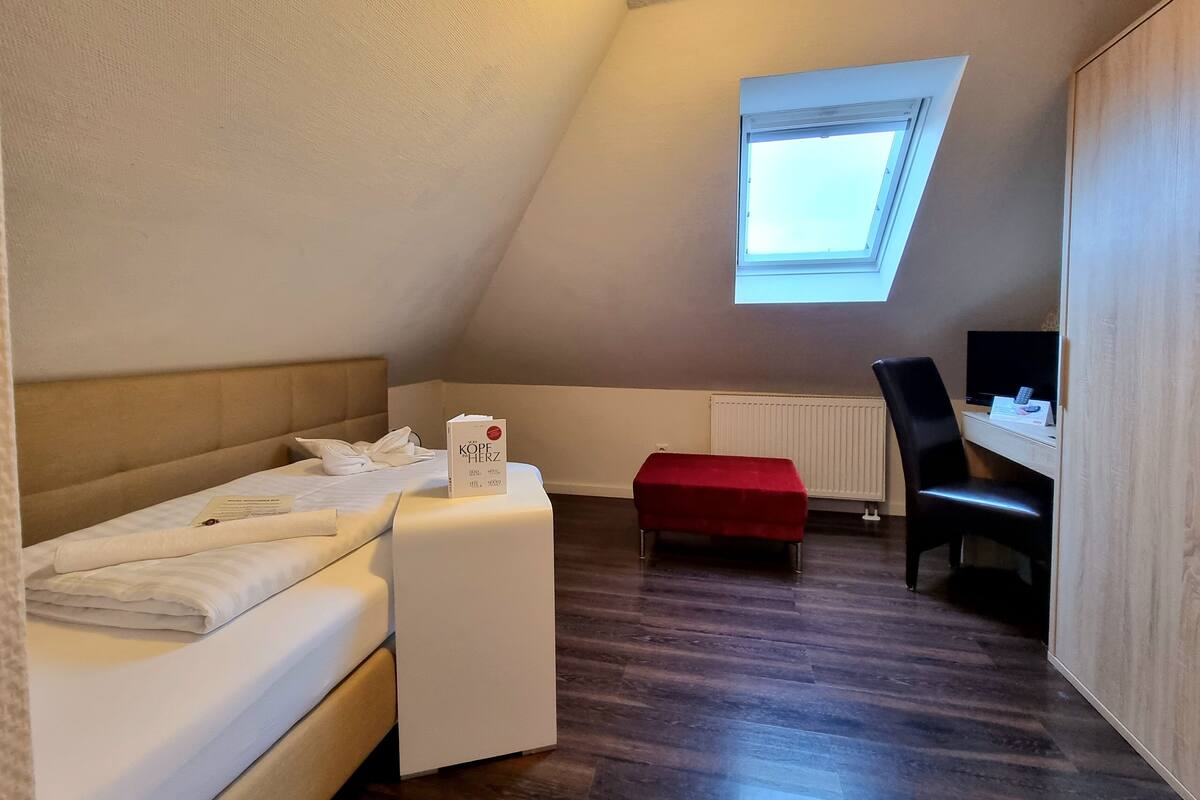 Quarto pequeno e moderno com uma cama, uma escrivaninha, uma cadeira, um banquinho vermelho e um claraboia.