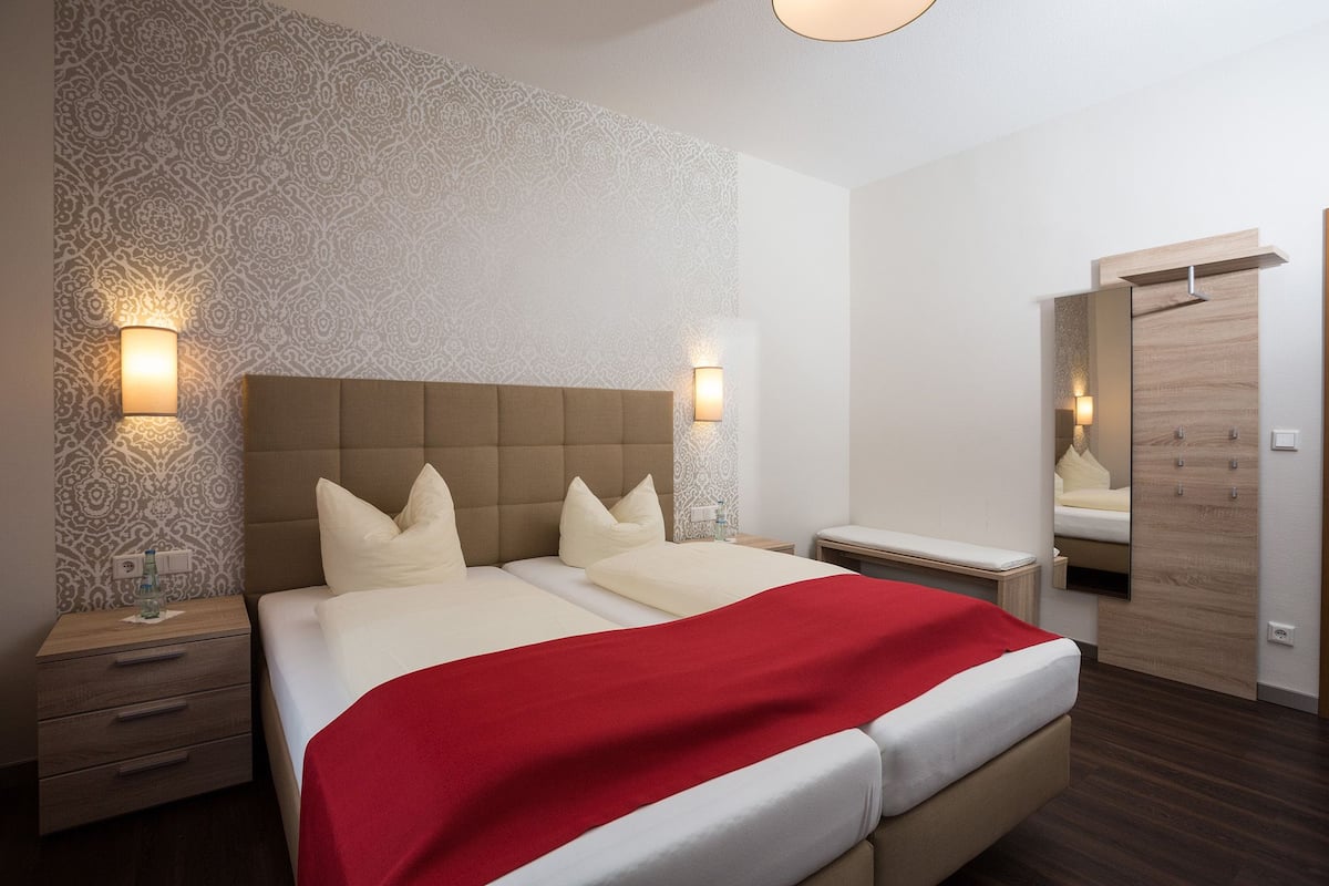 Quarto de hotel moderno com uma cama grande, duas mesas de cabeceira, um criado-mudo, um espelho e um guarda-roupa de madeira.