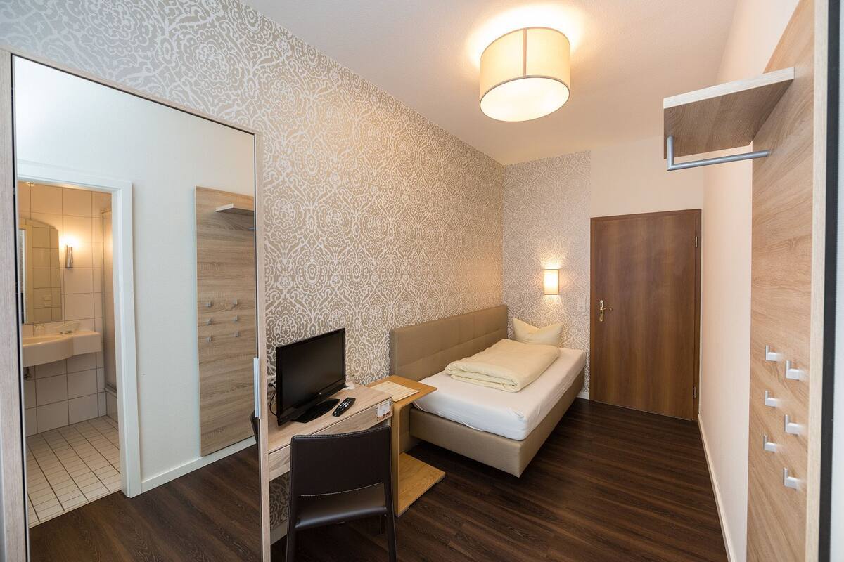 Quarto com cama, escrivaninha com computador, televisão, luminária na parede e porta.