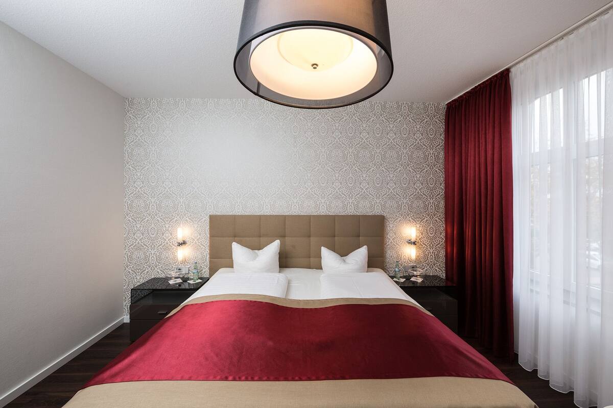 Quarto de hotel com uma cama grande, duas mesas de cabeceira, um cobertor vermelho, papel de parede estampado e uma janela ampla com cortinas.