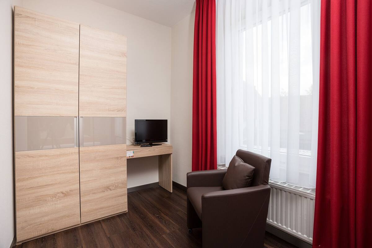 Quarto com guarda-roupa de madeira, escrivaninha com computador, poltrona de couro marrom e janela com cortinas vermelhas.