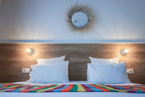 Cama bem arrumada com almofadas brancas e um cobertor listrado colorido, cabeceira de madeira e espelho redondo fixado na parede com design em formato de sol.