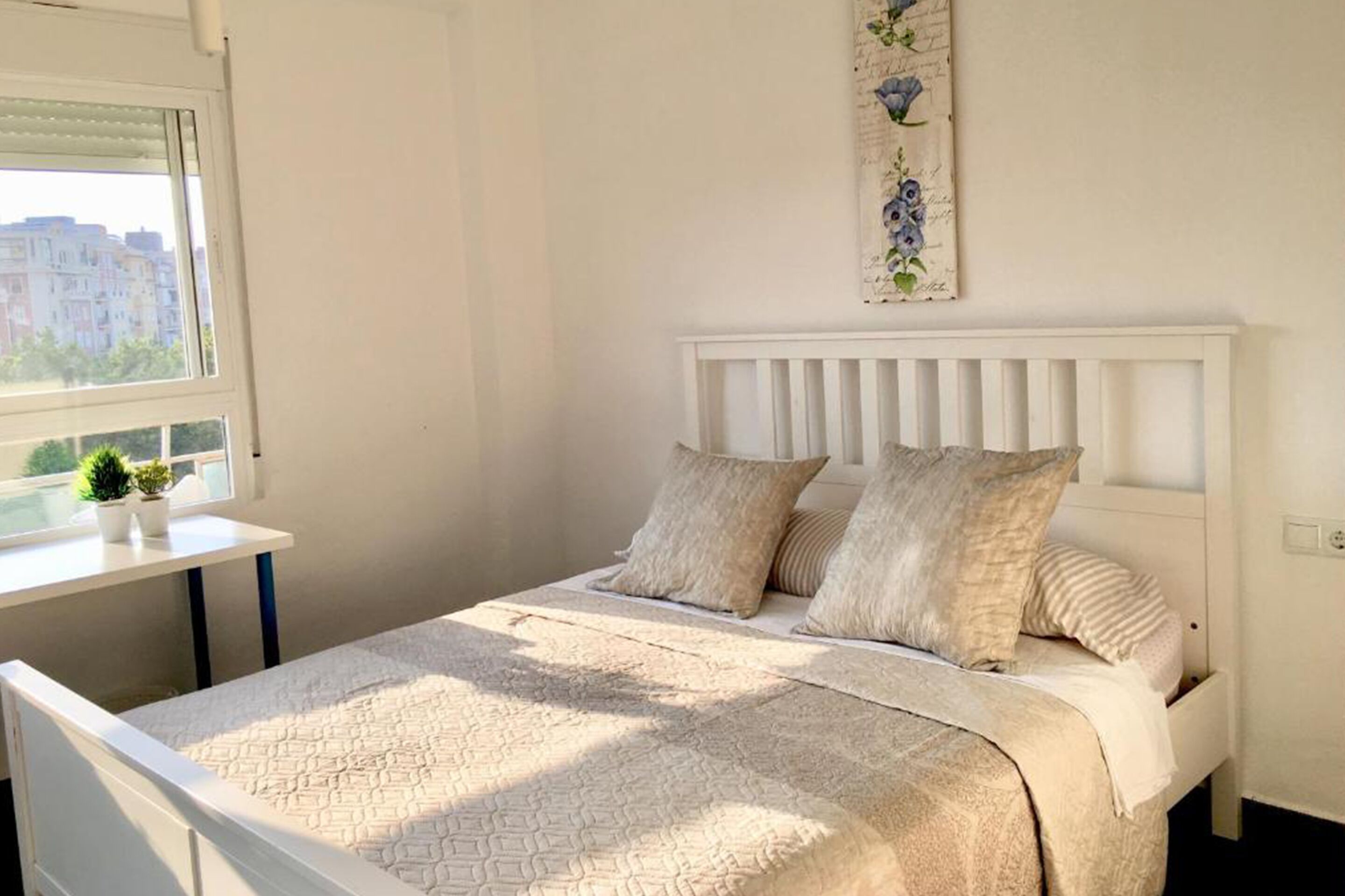Quarto com cama, janela, uma mesinha com planta e um objeto decorativo na parede.