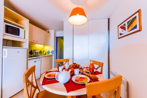 Uma área de refeições com mesa posta para dois, micro-ondas, cafeteira e uma geladeira ao fundo.