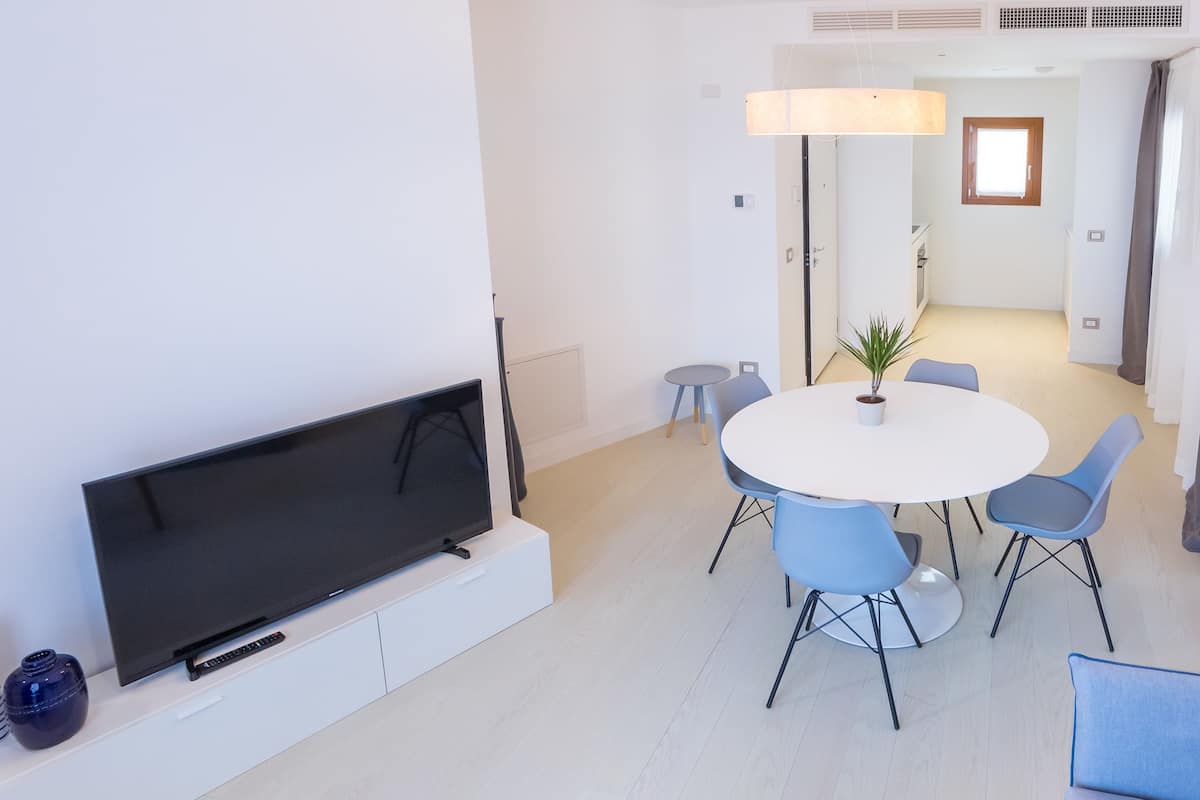 Uma sala de estar moderna com televisão de tela plana, uma mesa de jantar redonda com cadeiras azuis e uma pequena planta em vaso.