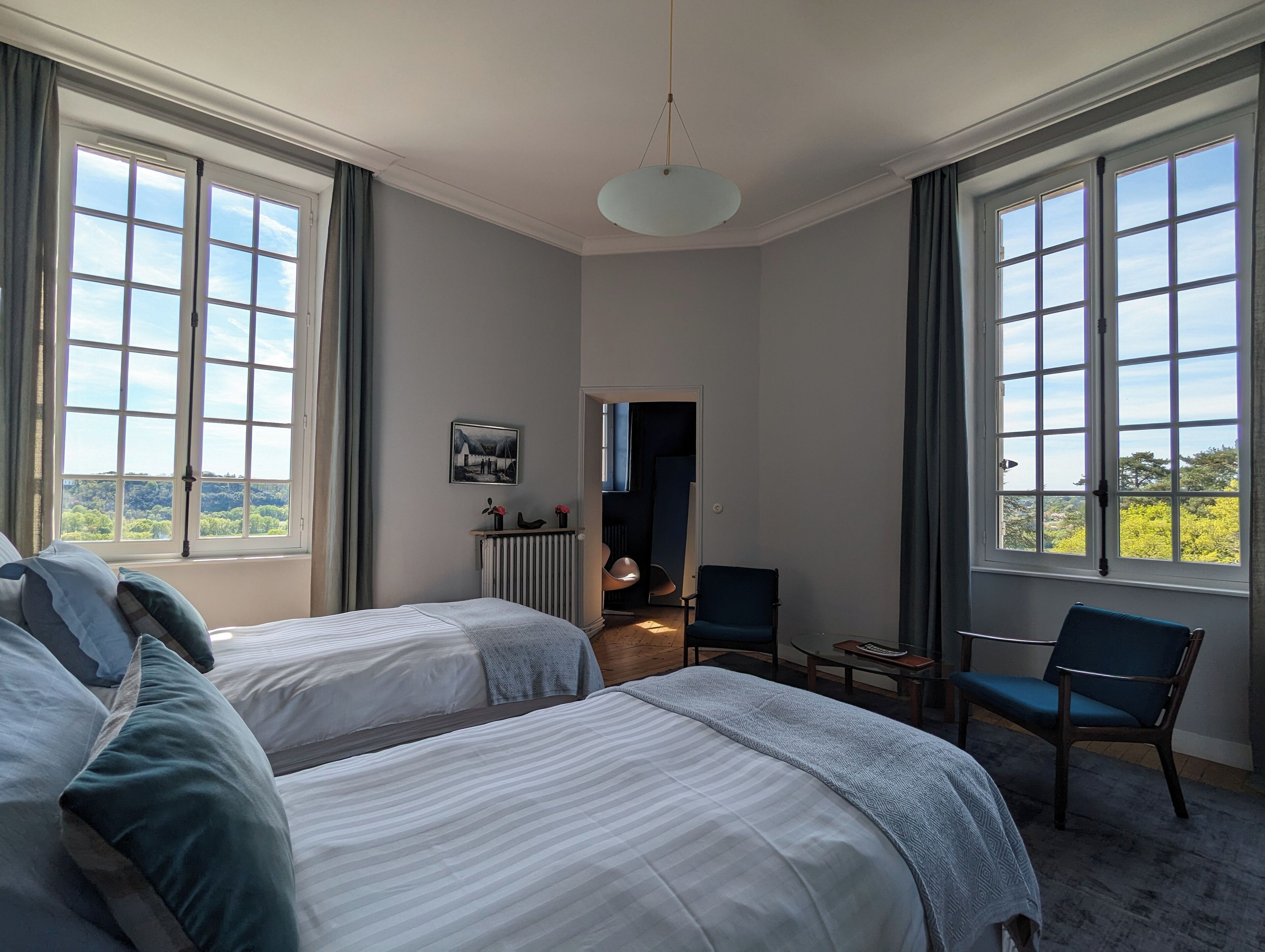 Quarto de hotel com duas camas, uma cadeira, uma mesinha e vista para o exterior.