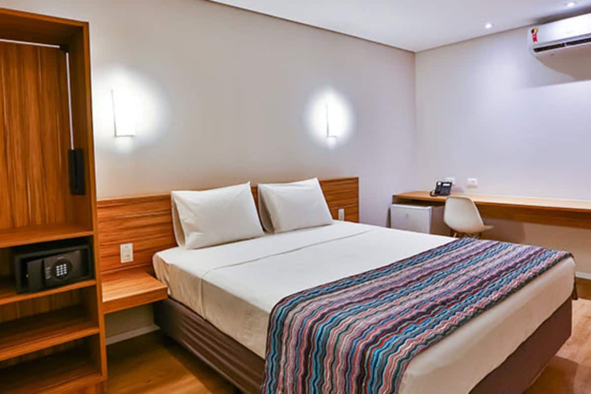 Quarto de hotel com cama, escrivaninha, cadeira, guarda-roupa e ar condicionado.
