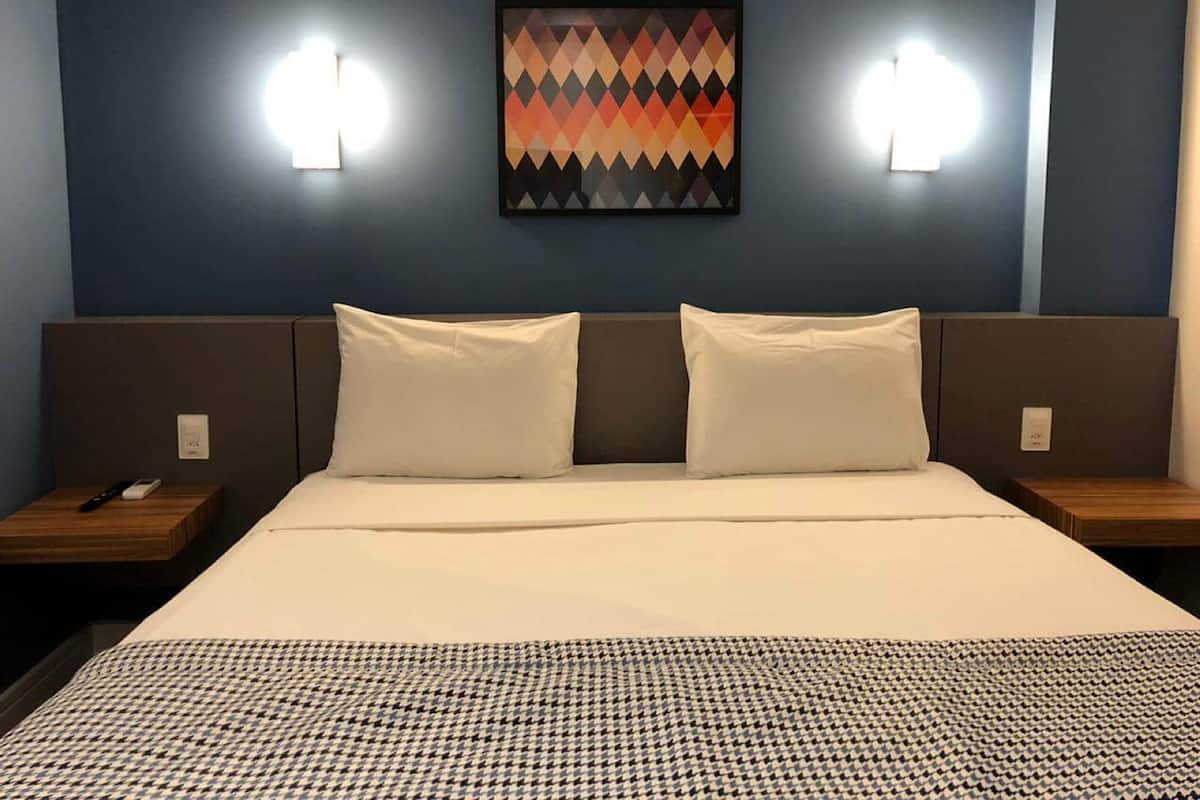 Quarto de hotel com uma cama grande, duas mesas de cabeceira, luminária fixada na parede e uma obra de arte emoldurada acima da cama.