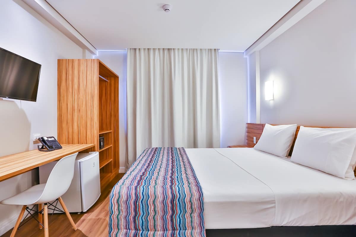 Quarto de hotel com cama, escrivaninha, cadeira, televisão e guarda-roupa.