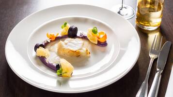 Um prato branco com um prato de peixe, decorado com detalhes roxos e amarelos, e um copo de vinho branco sobre uma mesa de madeira.
