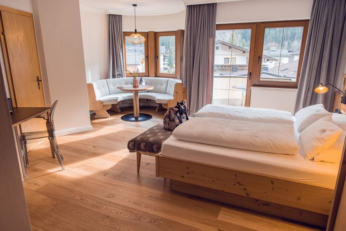 Quarto com cama de madeira, mesa redonda, sofá e janelas amplas.