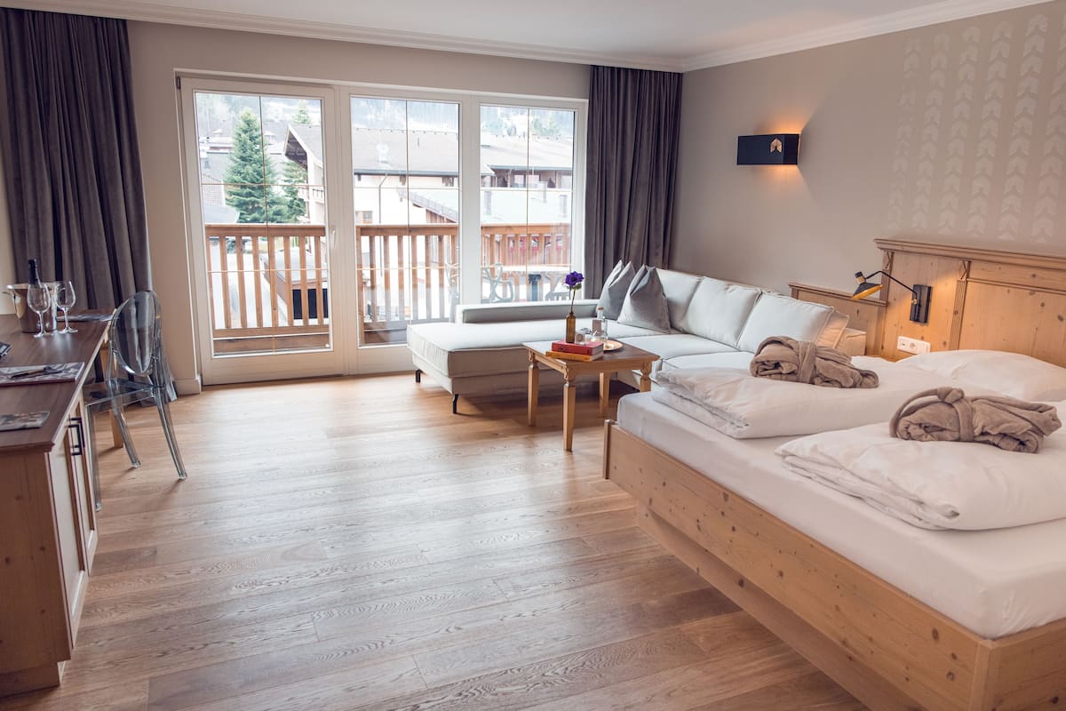 Quarto de hotel com piso de madeira, sofá, cama, mesa pequena e varanda com vista para uma árvore e prédios.