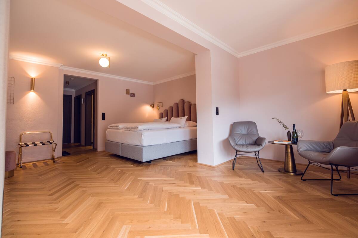 Quarto de hotel moderno com piso de madeira em padrão herringbone, uma cama, duas poltronas cinzas, uma mesinha redonda com abajur e luminária na parede.