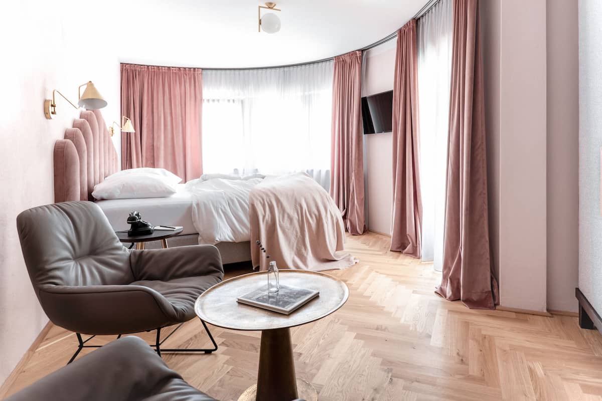 Quarto de hotel moderno com uma cama curva, um espaço de estar com uma poltrona e uma mesinha, e uma TV de tela plana fixada na parede.