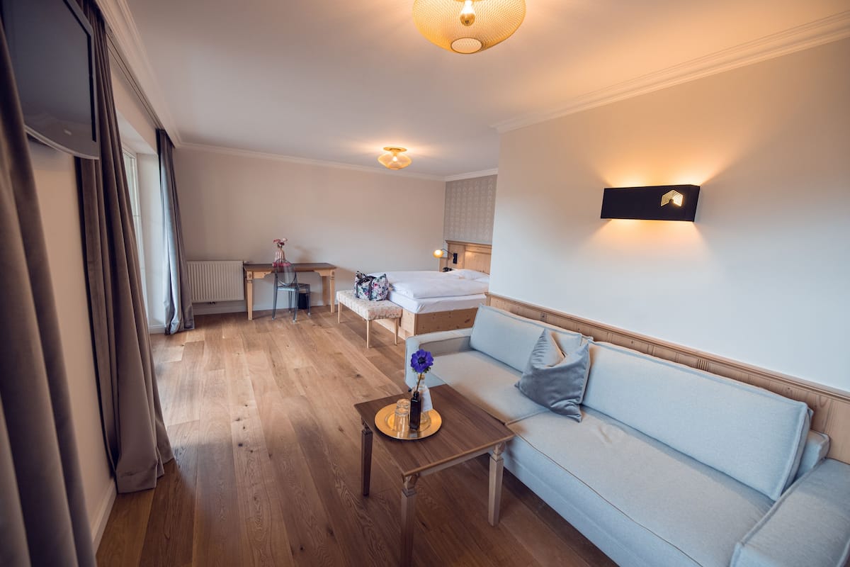 Quarto de hotel moderno com piso de madeira, sofá azul claro, pequena mesa de madeira com um vaso, cama com cabeceira branca e luminária fixada na parede.
