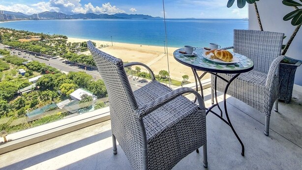Varanda com mobiliário de vime, uma mesa com um croissant e vista para a praia e o horizonte urbano.