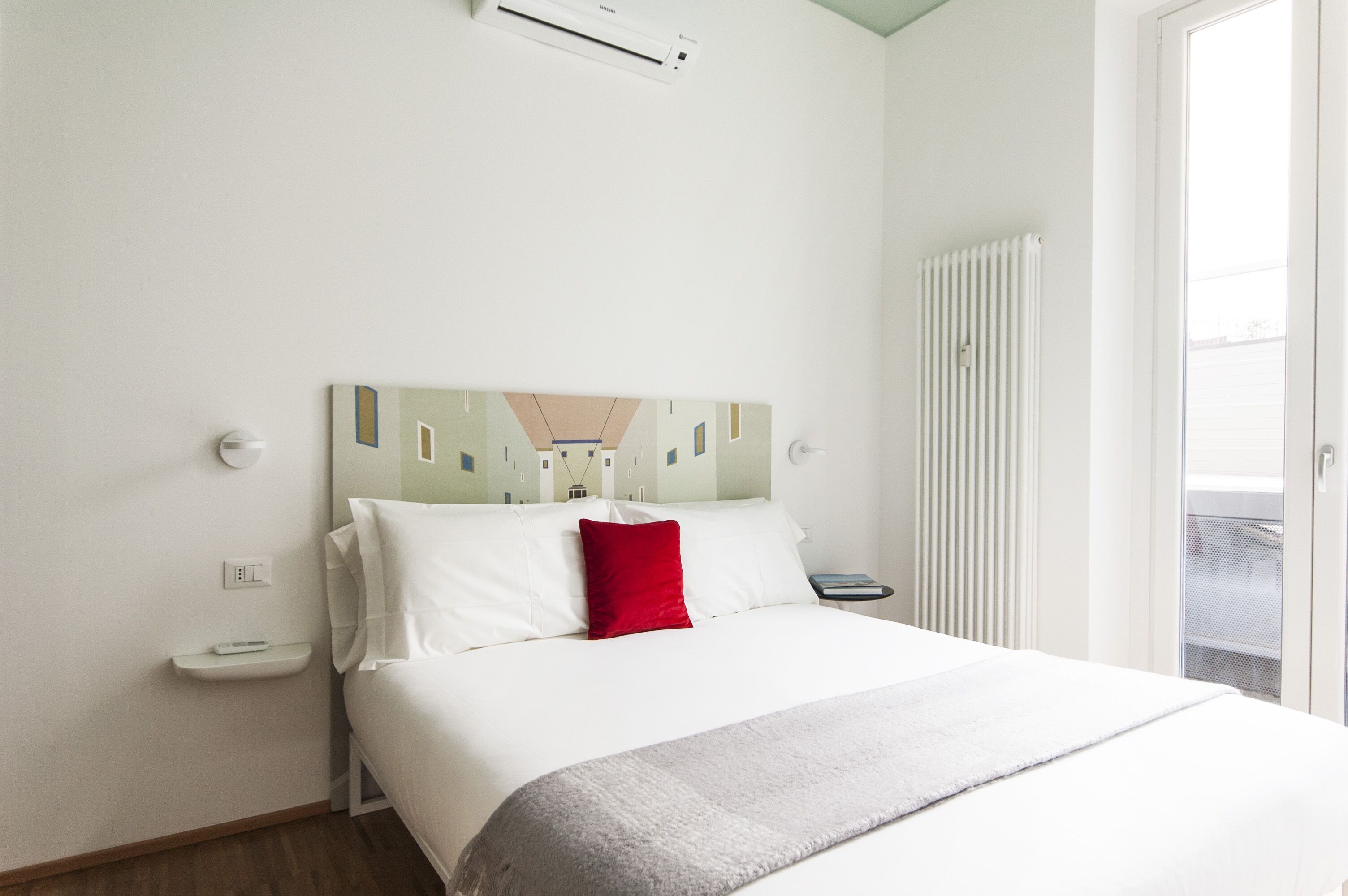 Quarto de hotel moderno com uma cama grande, roupa de cama branca, um travesseiro de destaque vermelho, uma mesinha de cabeceira com um controle remoto e um radiador.