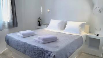 Quarto moderno com uma cama grande, roupa de cama branca, mesa de cabeceira com abajur e uma pequena planta.