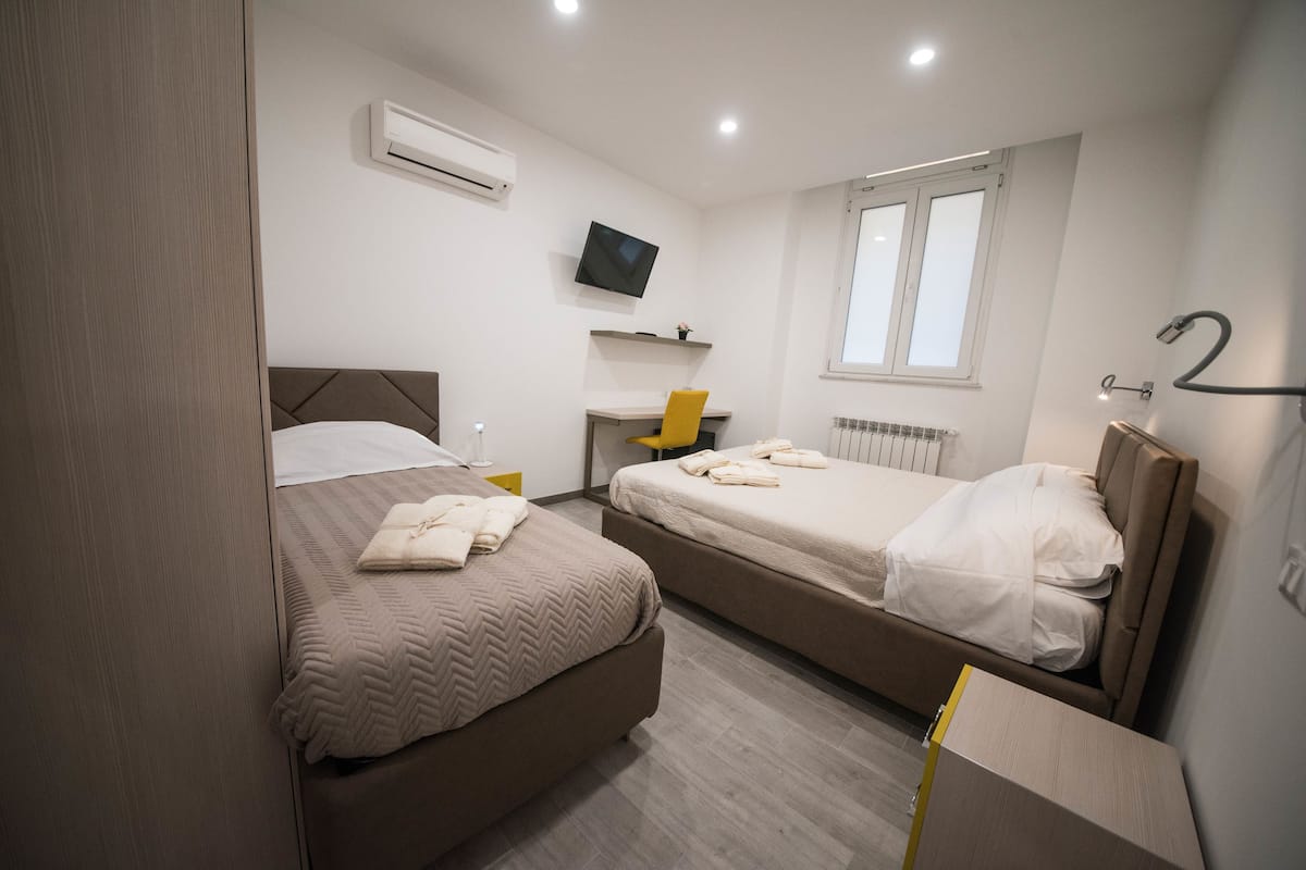 Quarto de hotel com duas camas, uma escrivaninha, uma cadeira, uma televisão e uma janela.