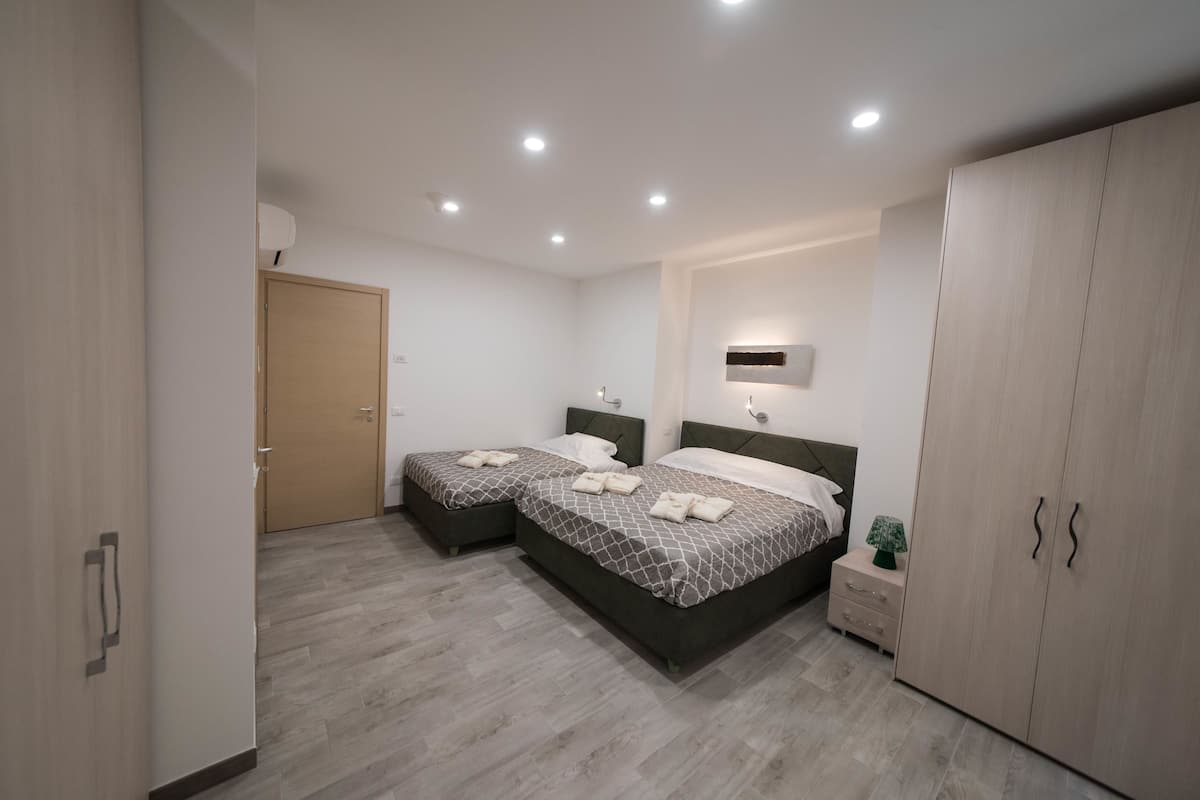Quarto com duas camas, um guarda-roupa, uma mesa de cabeceira e um candeeiro fixo na parede.