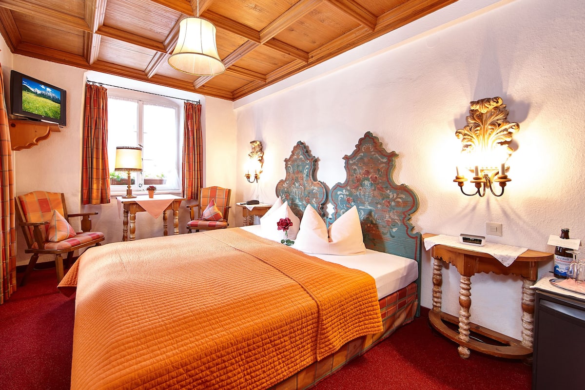 Quarto de hotel com uma cama grande, cabeceira de madeira, televisão, mesa com abajur e uma cadeira.