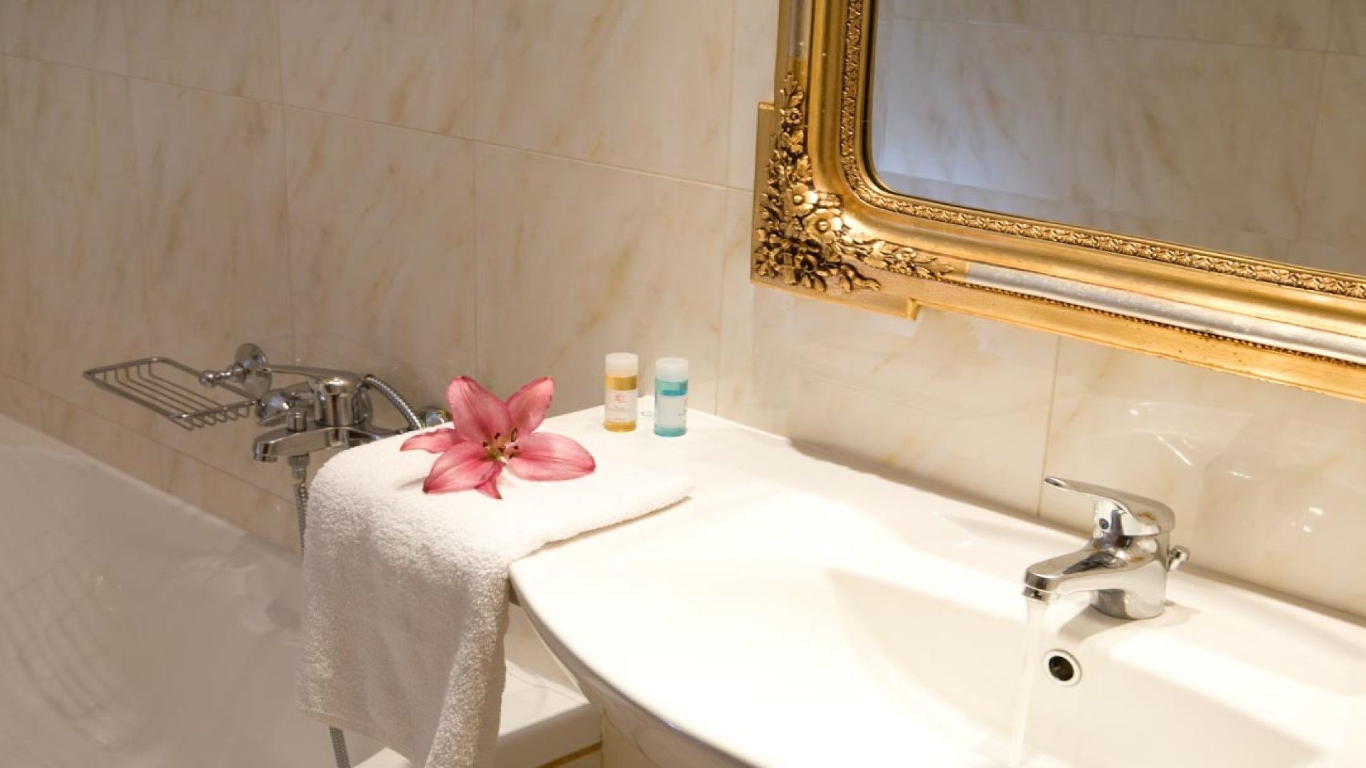 Casa de banho com lavatório branco, espelho com moldura dourada, toalha e uma flor rosa no balcão.