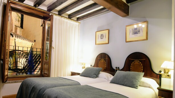 Quarto de hotel com duas camas, cabeceira de madeira, janela com vista para uma escadaria e quadros nas paredes.