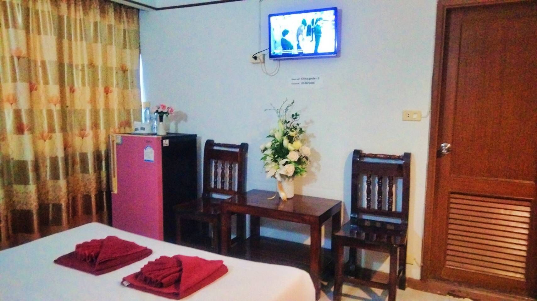 Quarto de hotel com uma cama, televisão na parede, uma mesinha com um vaso de flores, duas cadeiras e uma porta.
