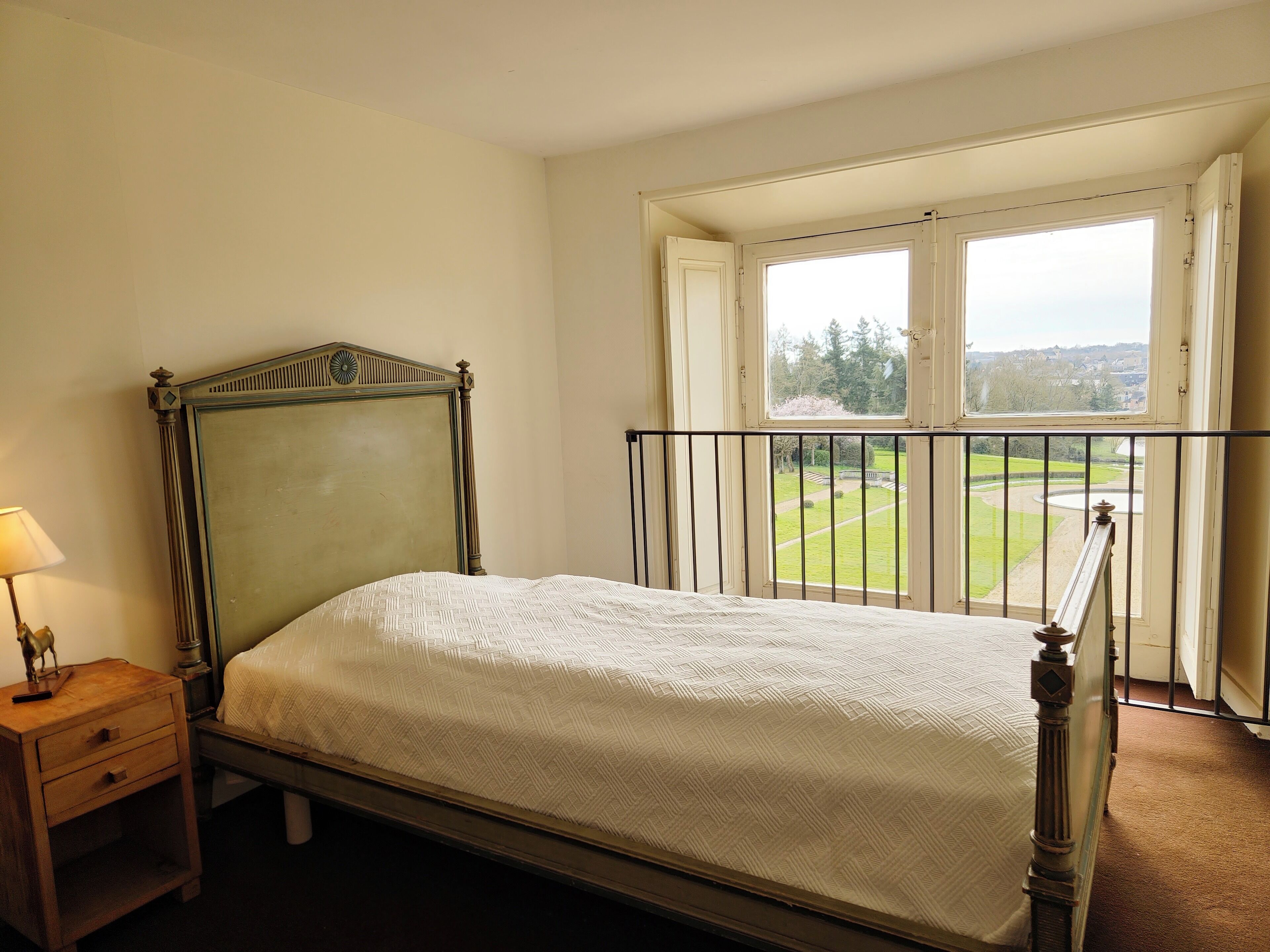 Quarto com cama, mesa de cabeceira, janela com vista para um jardim e guarda-corpo de varanda.