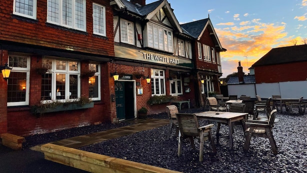 Um pub tradicional com uma placa que diz "The White Hart". Apresenta fachada de tijolos, várias janelas e área externa com mesas e cadeiras de madeira.