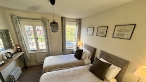 Quarto de hotel com duas camas, uma escrivaninha, um radiador e uma janela com cortinas.