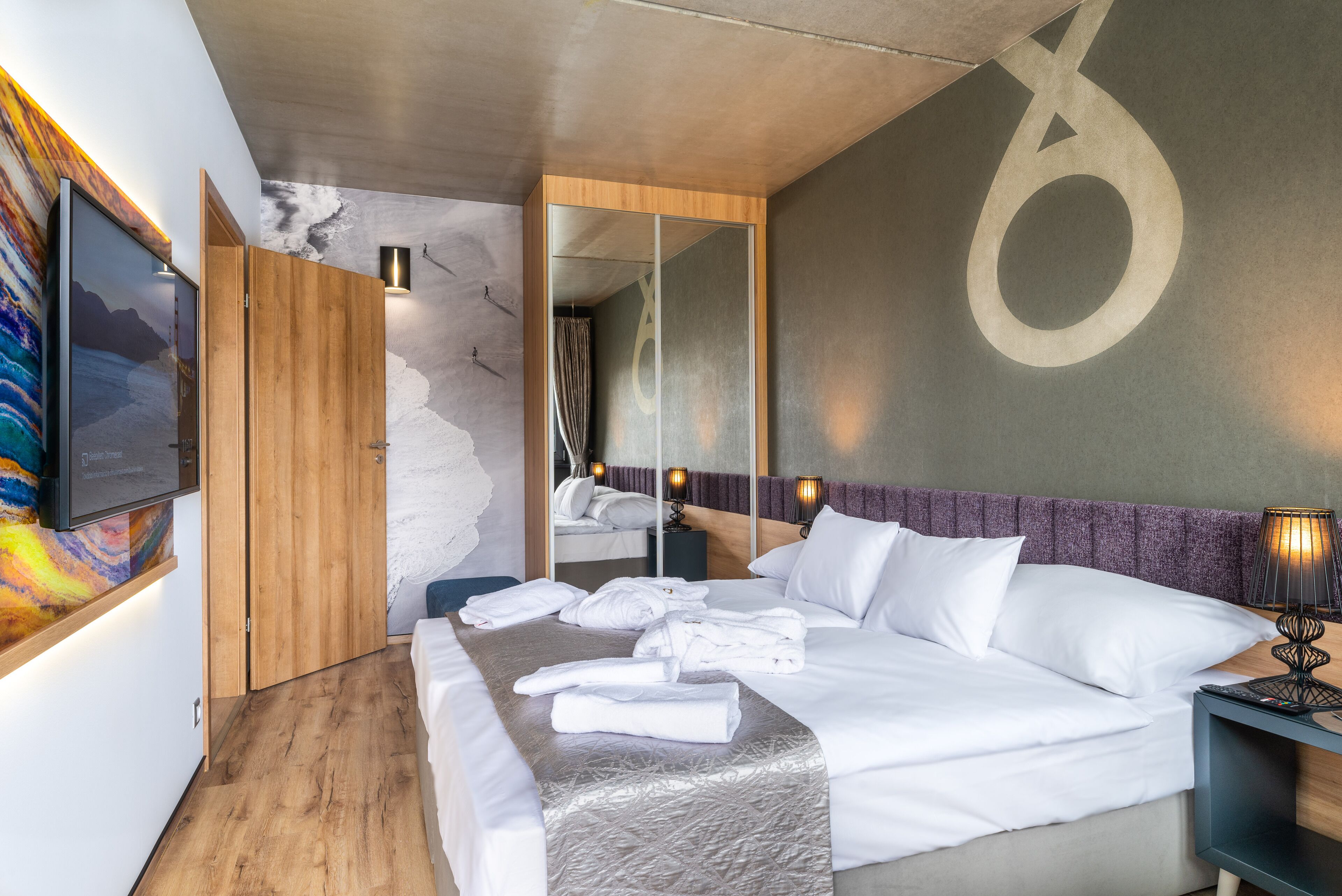 Quarto de hotel moderno com uma cama grande, TV fixada na parede, guarda-roupa de madeira e um espelho amplo.