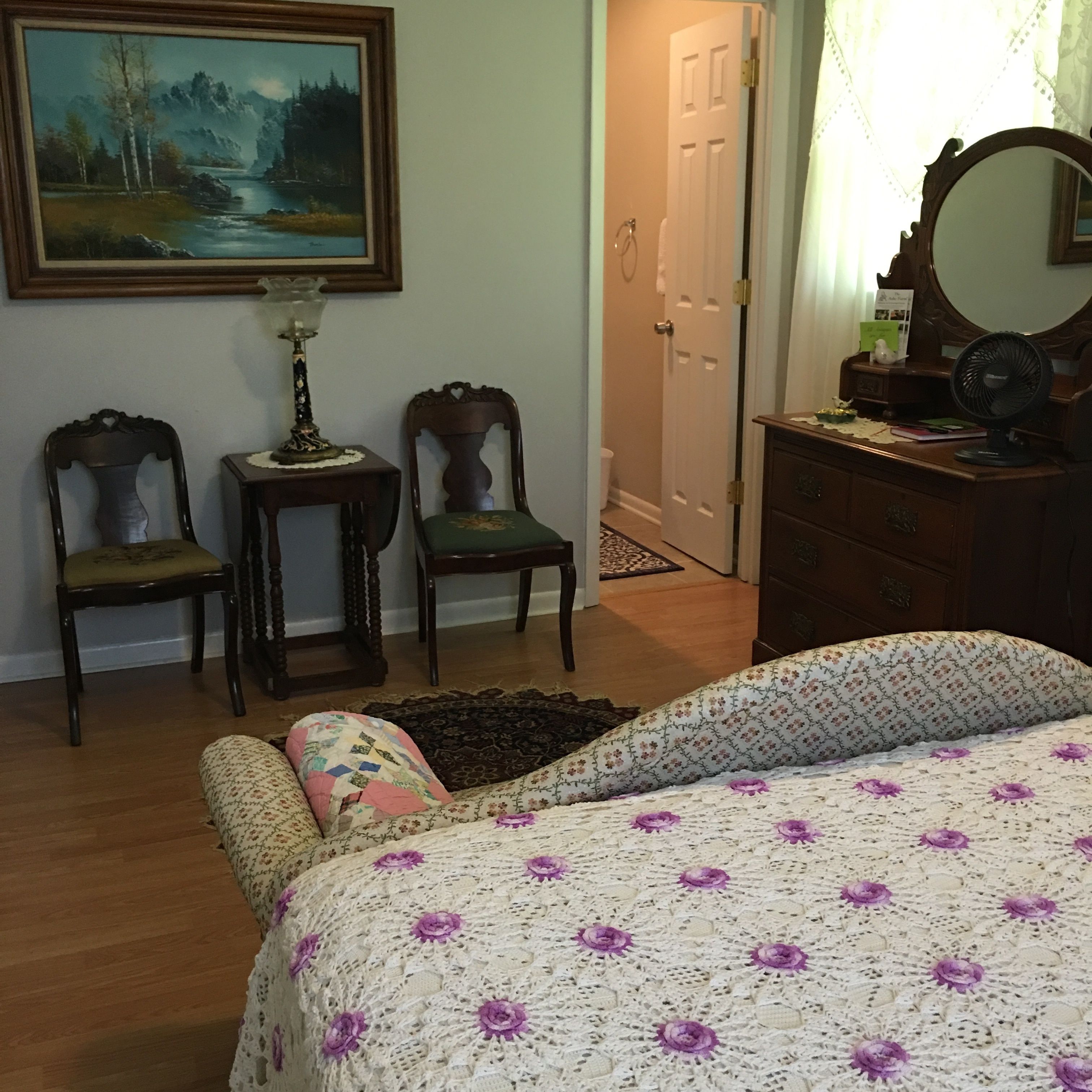 Quarto com uma cama, duas cadeiras, cômoda, espelho e pintura na parede.
