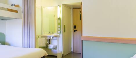 Quarto de hotel com uma cama de beliche, uma cama de solteiro, banheiro com pia e espelho, e uma porta que dá para outro quarto.