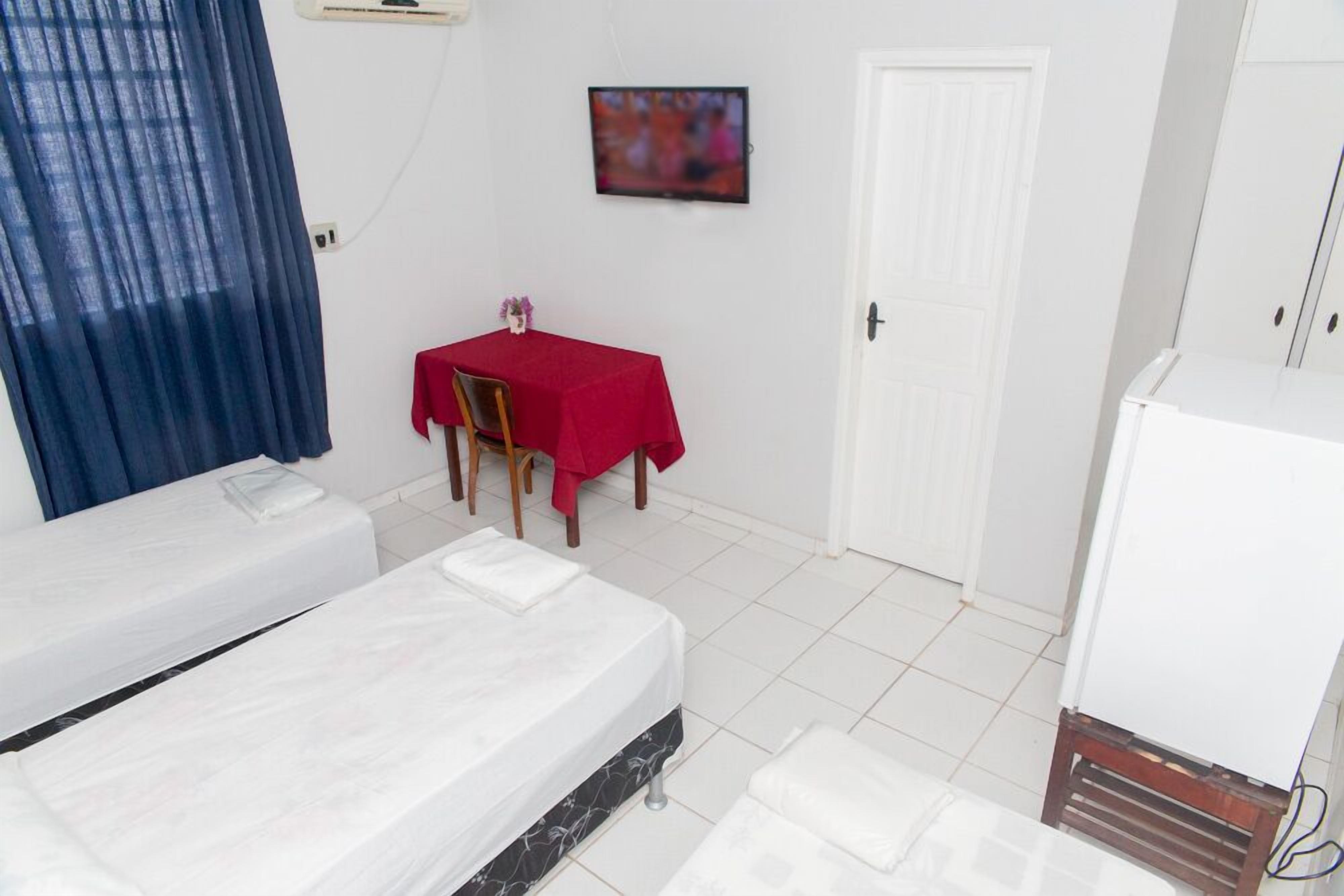 Quarto com duas camas, uma mesa pequena com toalha de mesa vermelha, televisão na parede e uma porta branca.