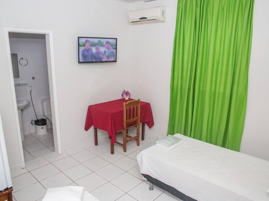 Quarto com cama, uma mesinha com cadeira, televisão na parede e banheiro visível através de uma porta aberta.