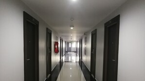 Um corredor comprido e estreito com portas em ambos os lados, um extintor fixado na parede e um chão limpo e espelhado.
