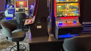 Uma fileira de máquinas caça-níqueis com um letreiro 'Dragon Cash', uma máquina 'Golden Century' e um letreiro 'Luxury Line'. Há dois banquetas pretas em frente às máquinas.