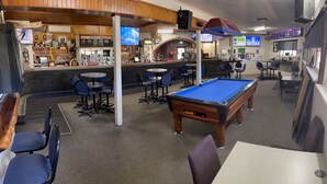 Uma sala de jogos com mesas de bilhar, cadeiras e um bar com televisão.