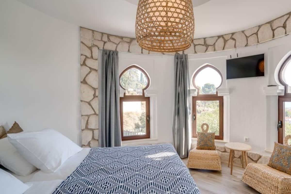 Quarto com cama, uma mesinha, duas cadeiras e TV fixada na parede.