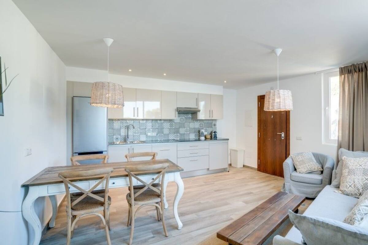 Uma cozinha moderna com armários brancos, uma mesa de jantar com cadeiras e um sofá com almofadas estampadas.