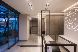 Um saguão de hotel moderno, com uma recepção de vidro, um grande relógio digital na parede e vista para o exterior através de amplas janelas.