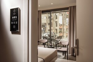 Quarto de hotel com porta de correr para uma varanda, uma cama, duas cadeiras, uma mesinha com uma garrafa e um painel de controlo fixado na parede.